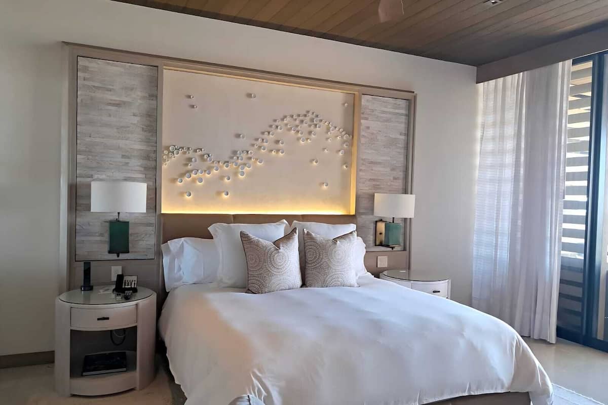 Quarto moderno com uma cama grande, duas mesas de cabeceira, um painel decorativo na parede e uma janela ampla com cortinas.