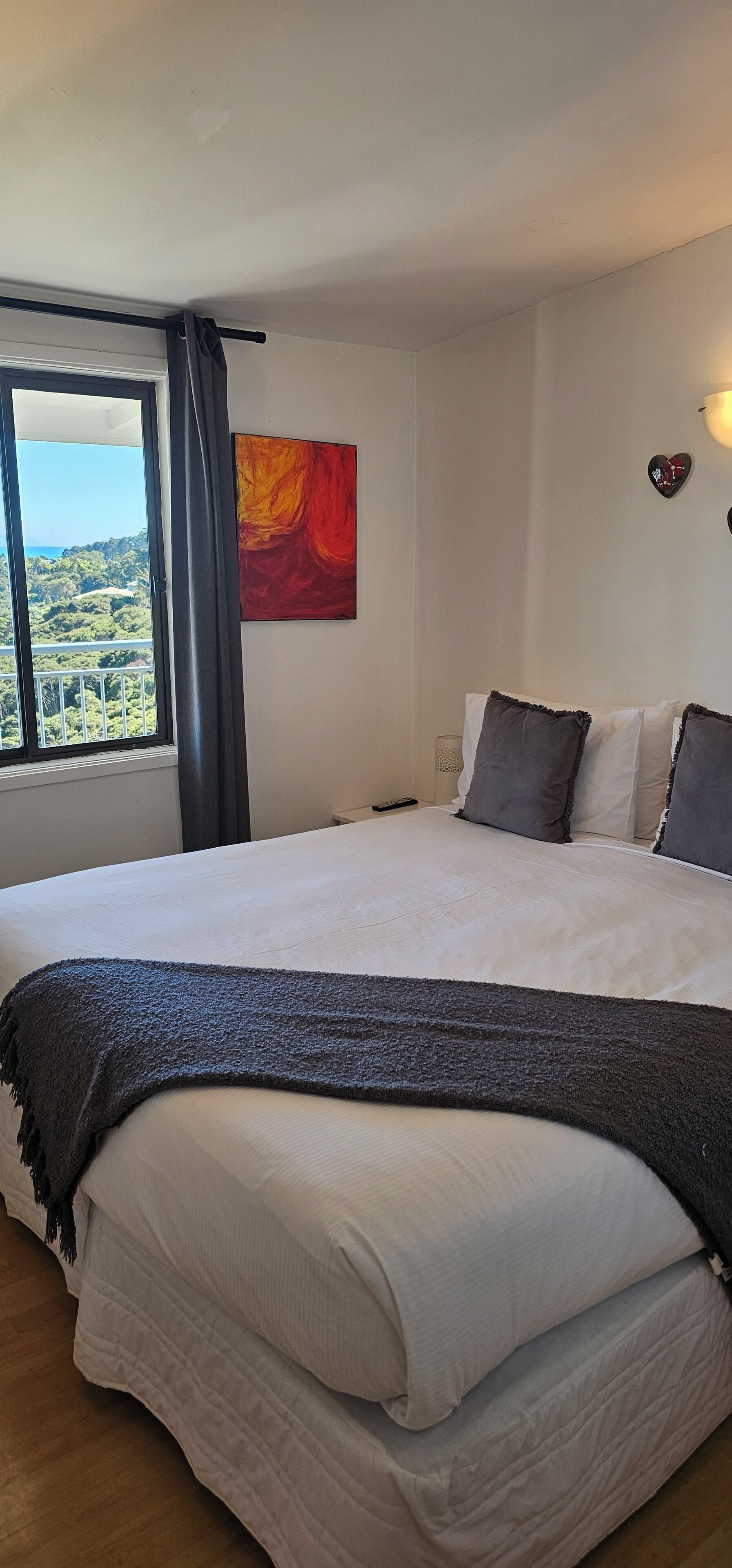 Quarto de hotel com uma cama grande, janela com cortinas, um quadro na parede e um lustre em formato de coração.