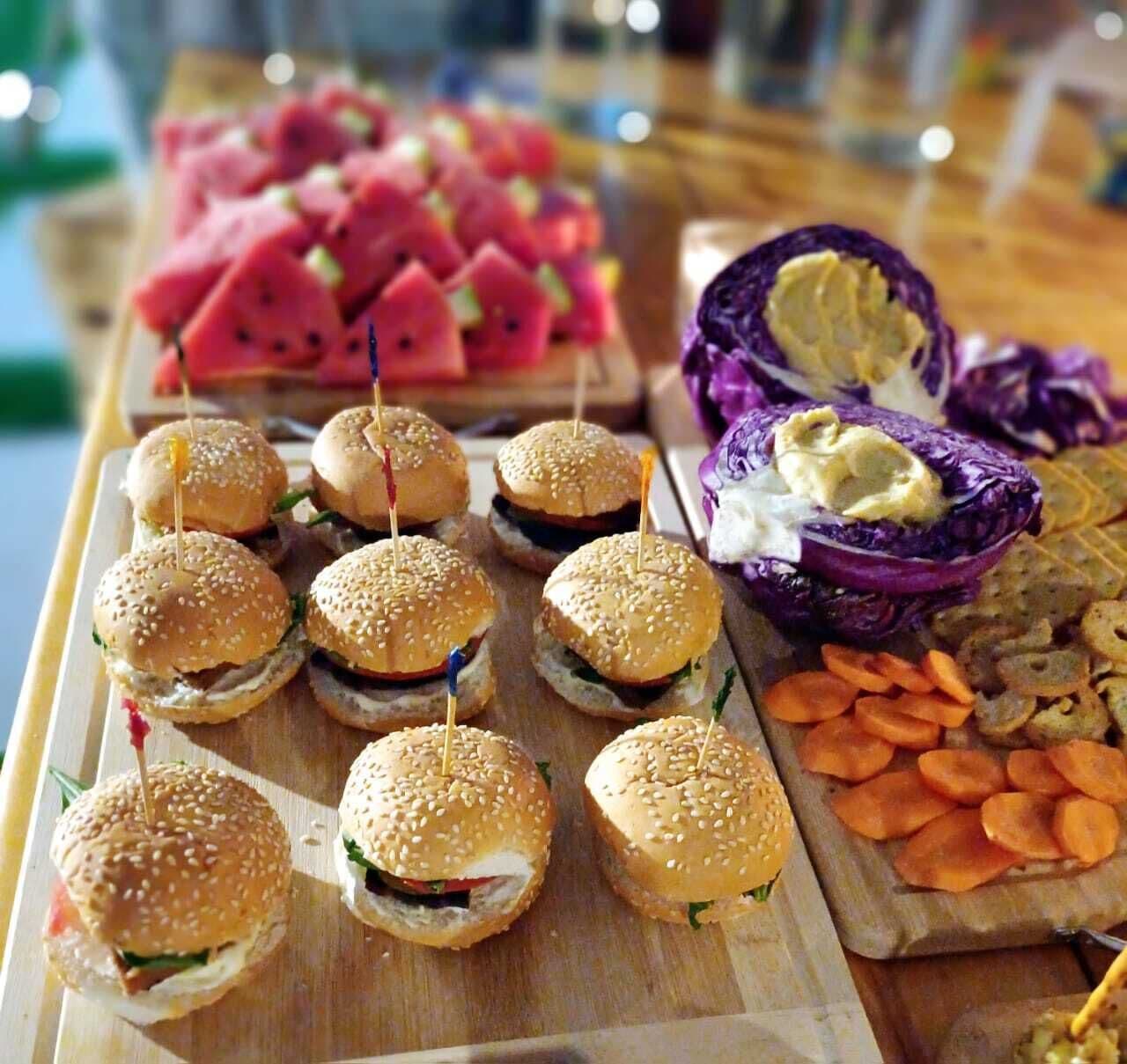 Uma mesa de buffet com mini sanduíches, fatias de melancia, uma salada com repolho roxo e palitos de cenoura.