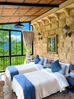 Quarto com duas camas, parede de pedra, janela com vista e ventilador de teto.
