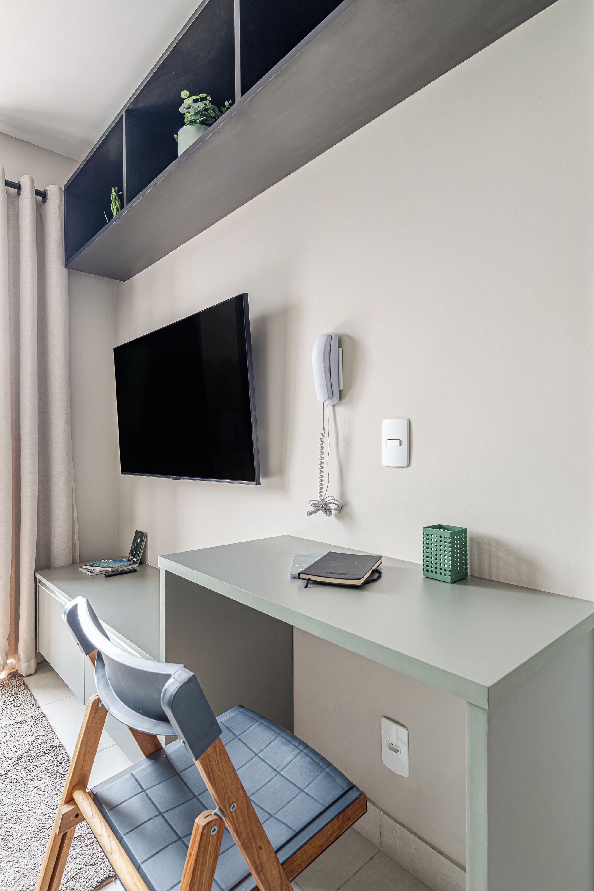 Quarto moderno e minimalista com televisão de tela plana na parede, escrivaninha com cadeira, telefone e uma pequena planta numa prateleira.