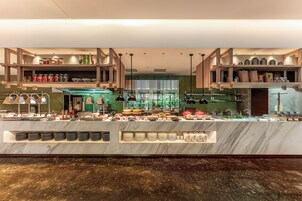 Um lobby moderno de hotel, com balcão de recepção em mármore, área de buffet e vista para o horizonte urbano através de amplas janelas.