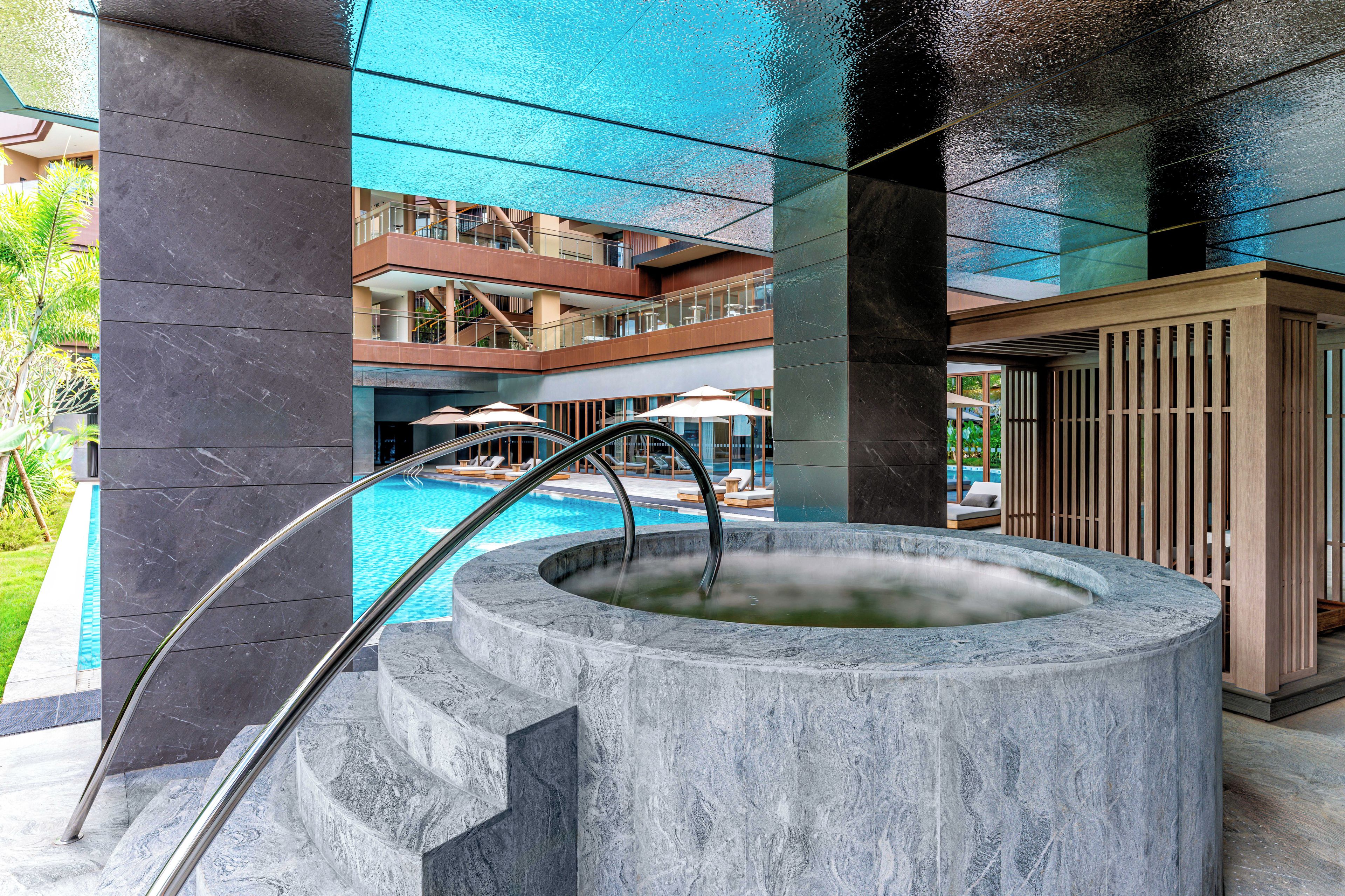 Uma área de spa com hidromassagem, piscina e vista para um edifício de hotel com vários andares e varandas.