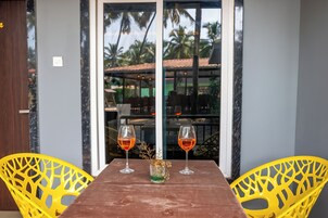 Uma mesa de madeira com dois copos de vinho tinto e um pequeno vaso com flores secas, ladeada por cadeiras amarelas, com vista para palmeiras e área de estar ao ar livre através de uma janela.