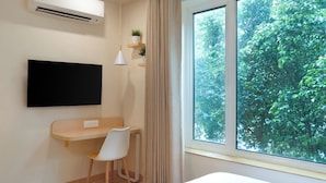 Quarto moderno com cama, escrivaninha com cadeira, TV na parede, ar condicionado e uma grande janela com vista para árvores.