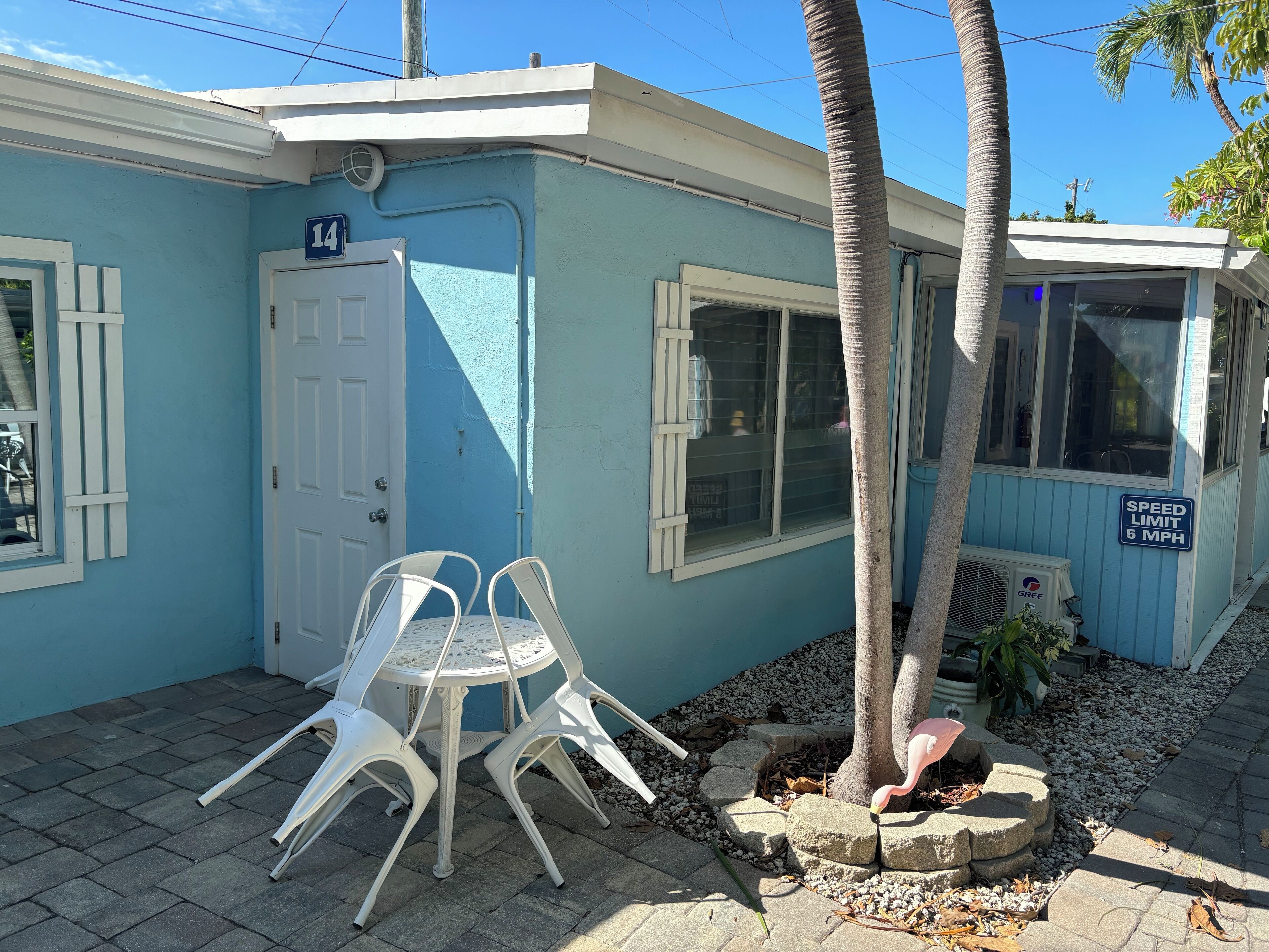 Um pequeno edifício térreo com fachada azul, porta branca e uma janela com placa de limite de velocidade. Cadeiras de plástico branco e uma pequena mesa estão do lado de fora, e uma árvore com uma estátua de flamingo por perto.