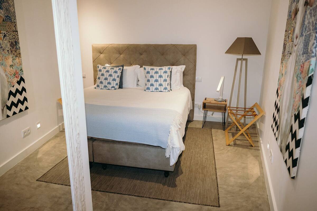 Um quarto com cabeceira estofada, cama com almofadas estampadas, luminária de cabeceira e uma cadeira de madeira.