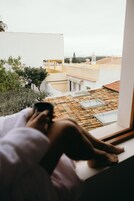 Pessoa sentada perto de uma janela com vista para um pátio com edifícios e árvores.