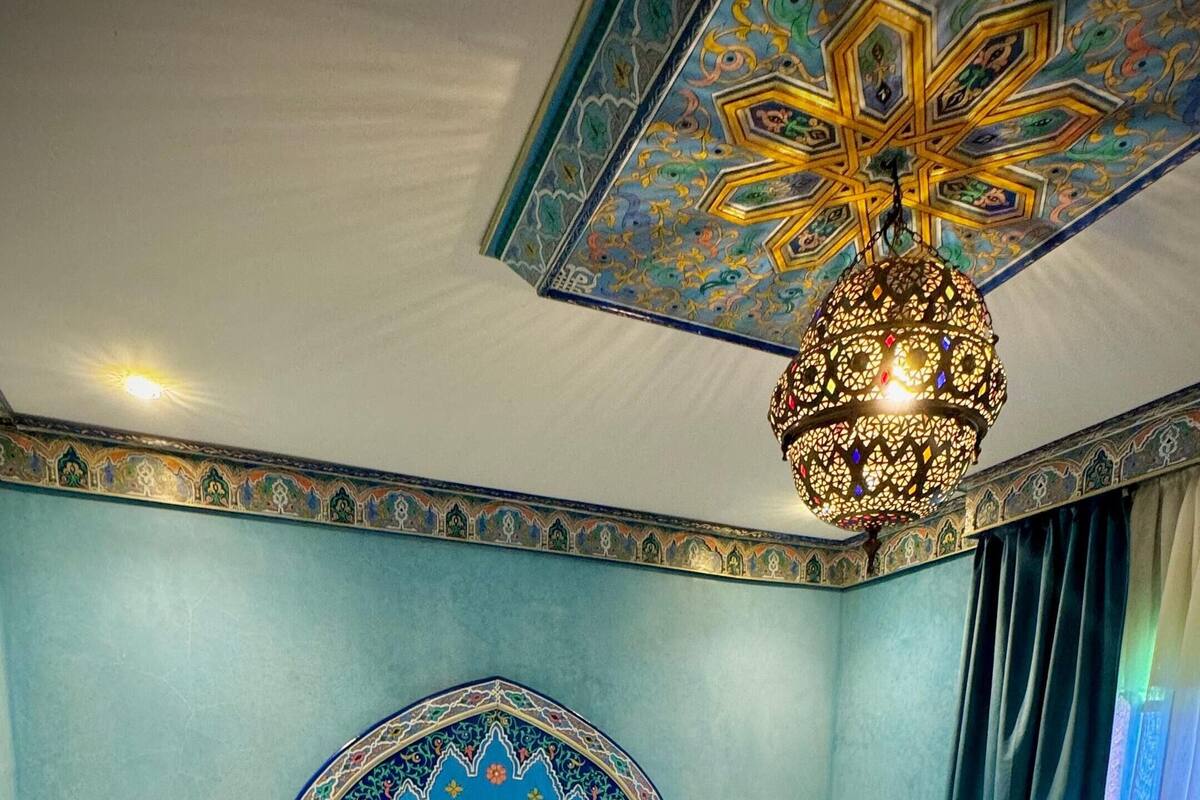 Quarto com cama grande, paredes com padrão azul, teto decorativo e luminária pendente.