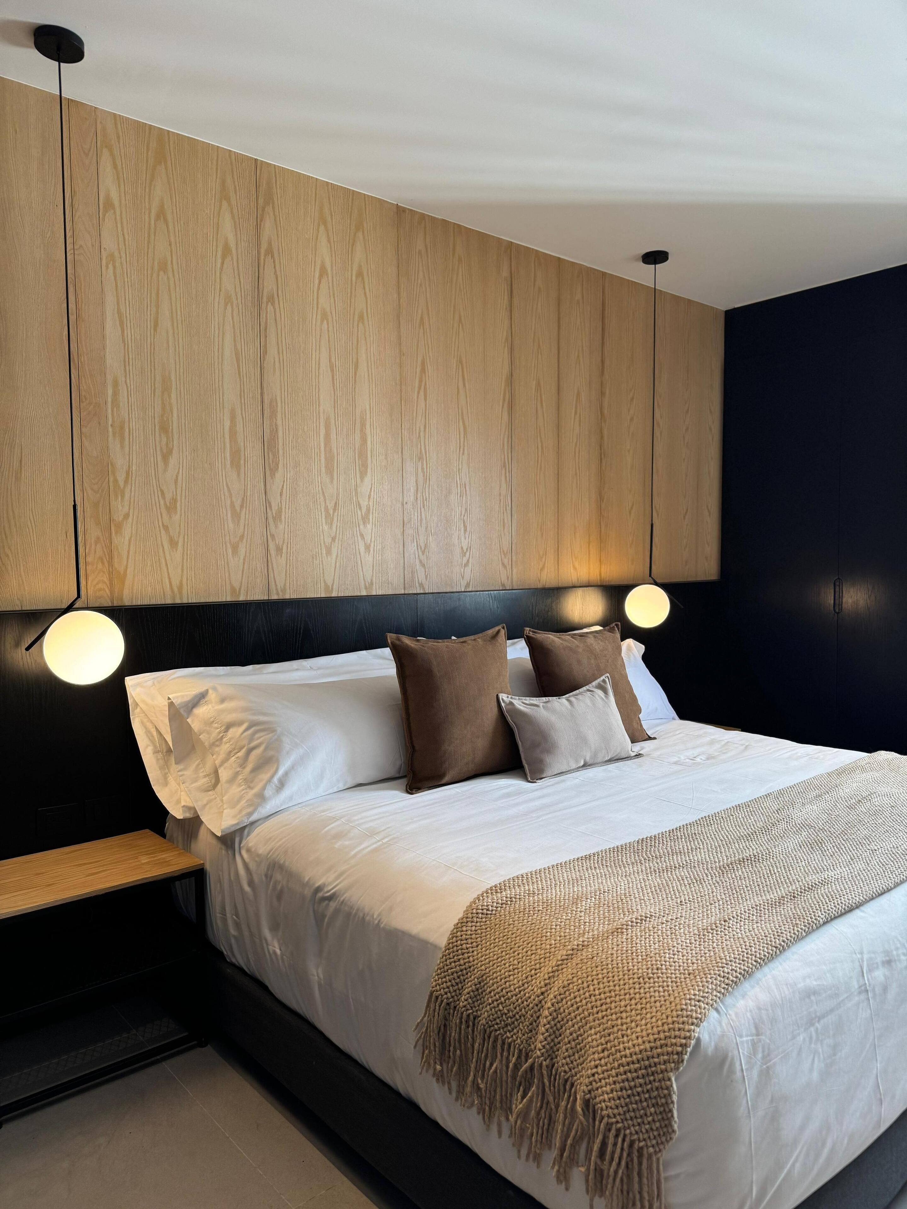 Quarto moderno com cabeceira de madeira, cama com roupa de cama branca e almofadas castanhas, mesa de cabeceira e duas luzes fixadas na parede.
