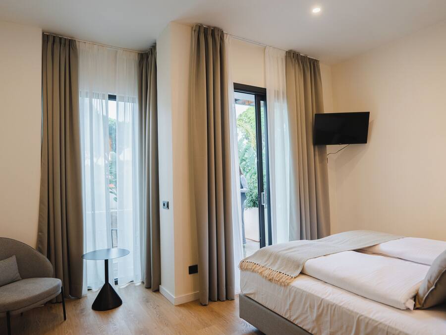 Quarto de hotel com uma cama, uma cadeira, uma mesinha e uma televisão presa à parede.