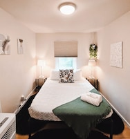 Quarto de hotel com cama, abajures, janela com persianas, planta pendente e obras de arte nas paredes.