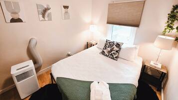 Quarto de hotel com cama, mesa de cabeceira, cadeira, janela com persianas e decoração na parede.