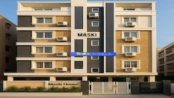 Um edifício de hotel de vários andares chamado "Maski House", com design arquitetônico moderno, com unidades de ar condicionado em cada andar e uma entrada com portão.