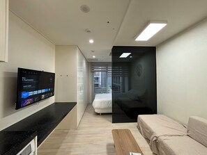 Quarto de hotel moderno com televisão de tela plana na parede, uma cama com cabeceira escura, um sofá de cor clara e um banco de madeira.
