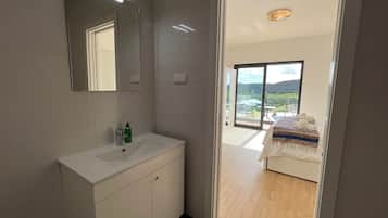 Casa de banho moderna com uma pia branca, espelho e armários. Uma porta de vidro deslizante dá acesso a um quarto com cama e vista para uma varanda.
