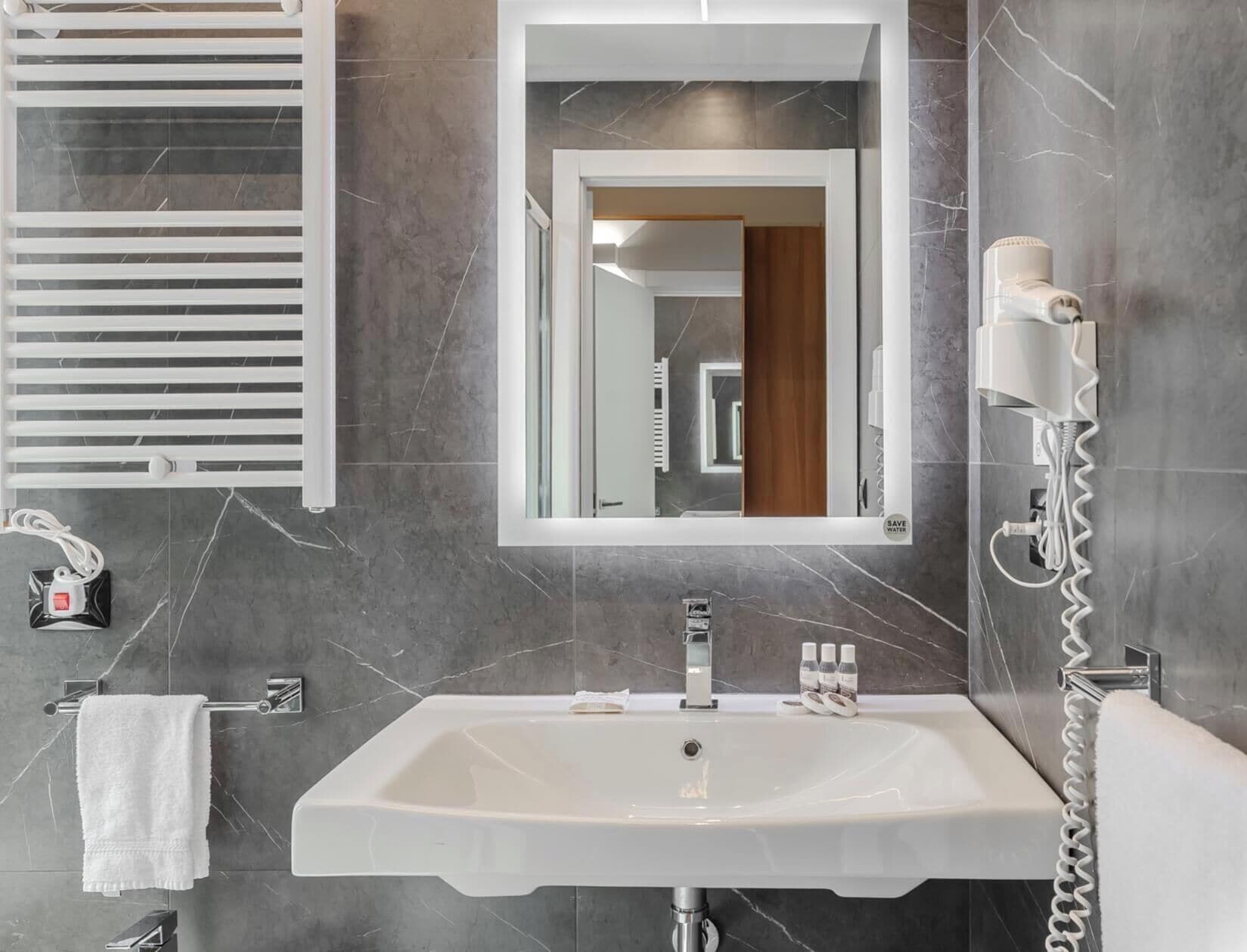 Casa de banho com lavatório branco, espelho, porta-toalhas e secador de cabelo fixado na parede.