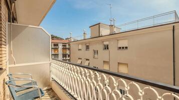 Uma varanda com grades de metal, uma cadeira e vista para edifícios próximos.