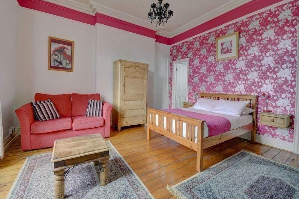 Quarto com cama de madeira, um sofá vermelho, cómoda de madeira e um candelabro.