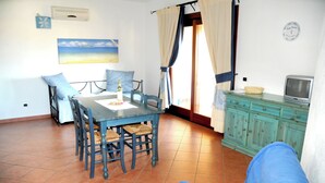 Uma sala de jantar com mesa e cadeiras, um sofá, uma televisão sobre um móvel e um quadro na parede.