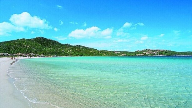 Uma praia com águas turquesa cristalinas, uma faixa de areia e um relevo montanhoso ao fundo.