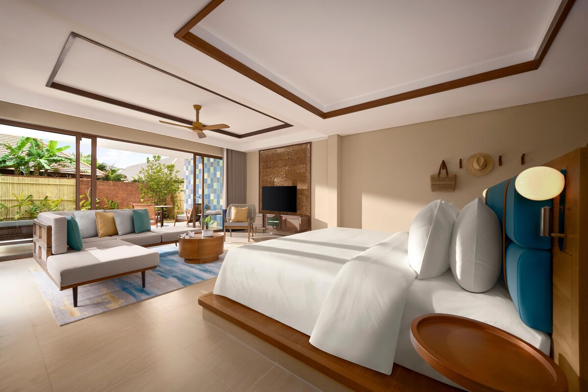 Quarto de hotel moderno com uma cama grande, um sofá, uma TV e uma varanda com plantas.