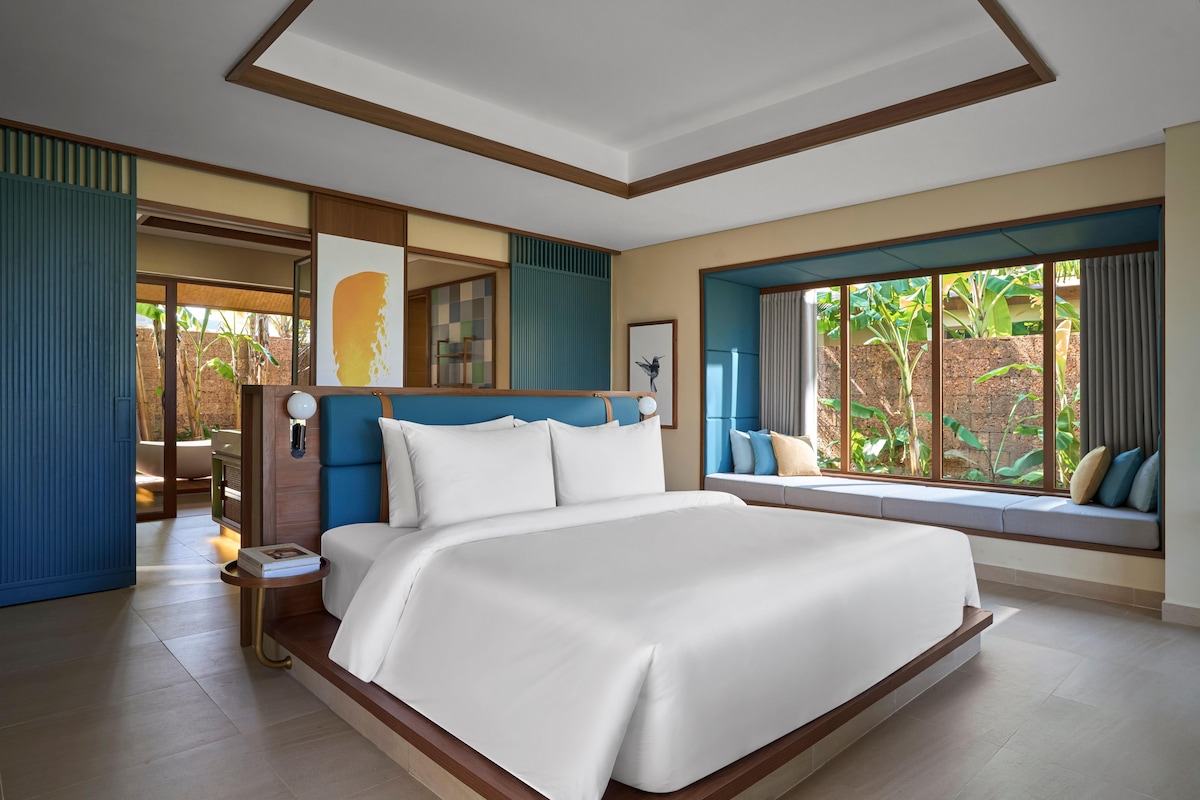 Quarto de hotel moderno com uma cama grande, assentos integrados junto à janela e vista para áreas verdes lá fora.