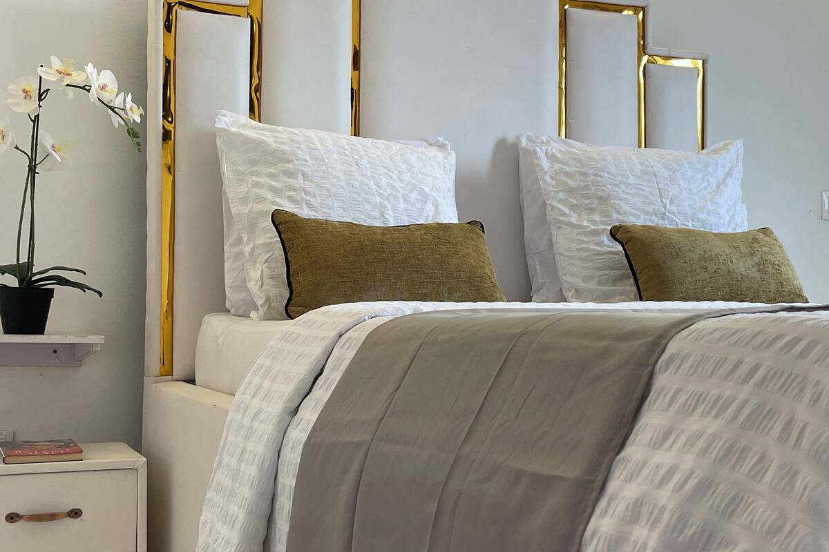Cama bem arrumada com roupa de cama branca e bege, duas cabeceiras com moldura dourada e uma planta na mesinha de cabeceira.