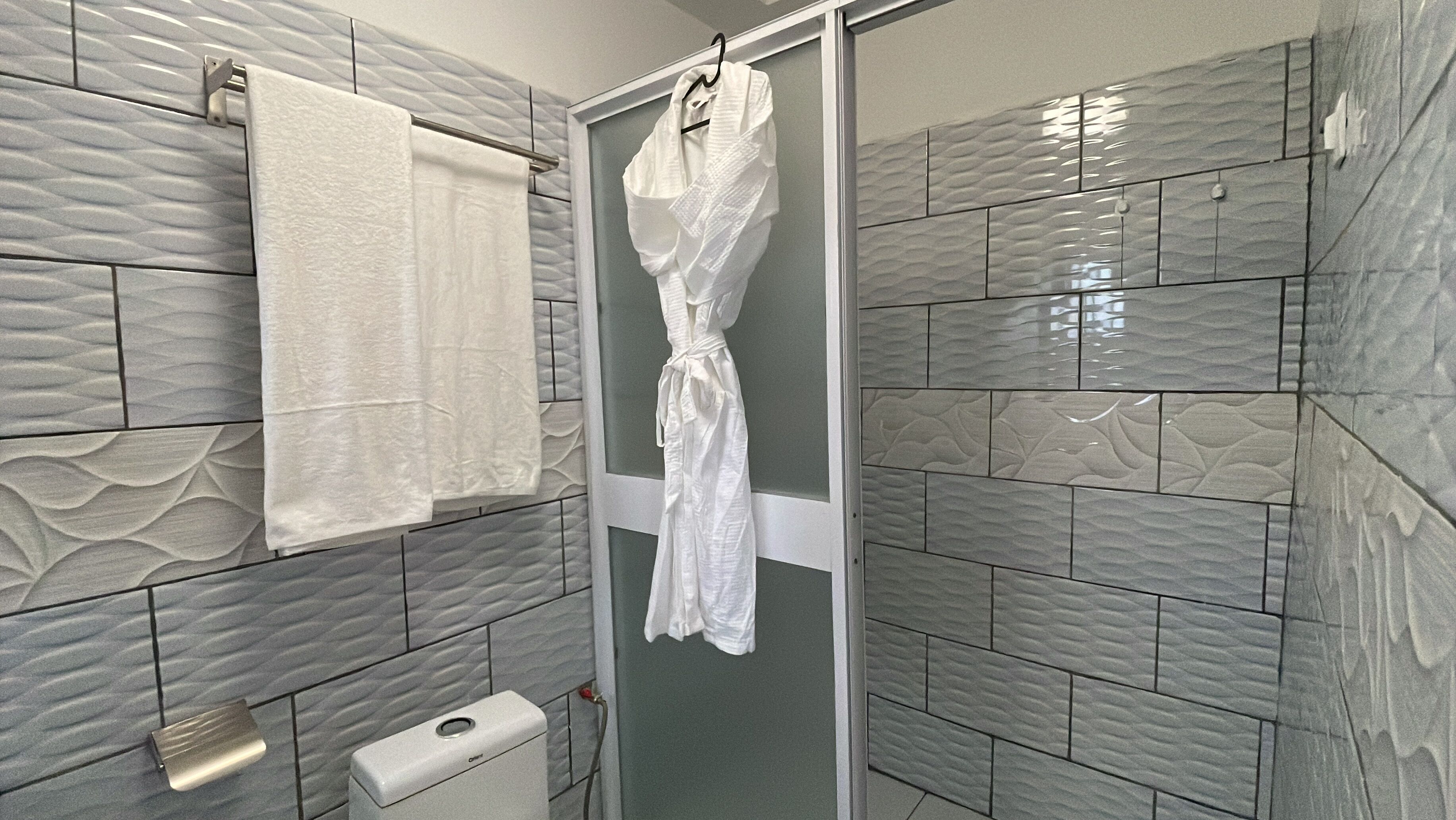 Casa de banho com azulejos cinzentos com textura, uma toalha branca num suporte e um roupão branco pendurado numa porta.