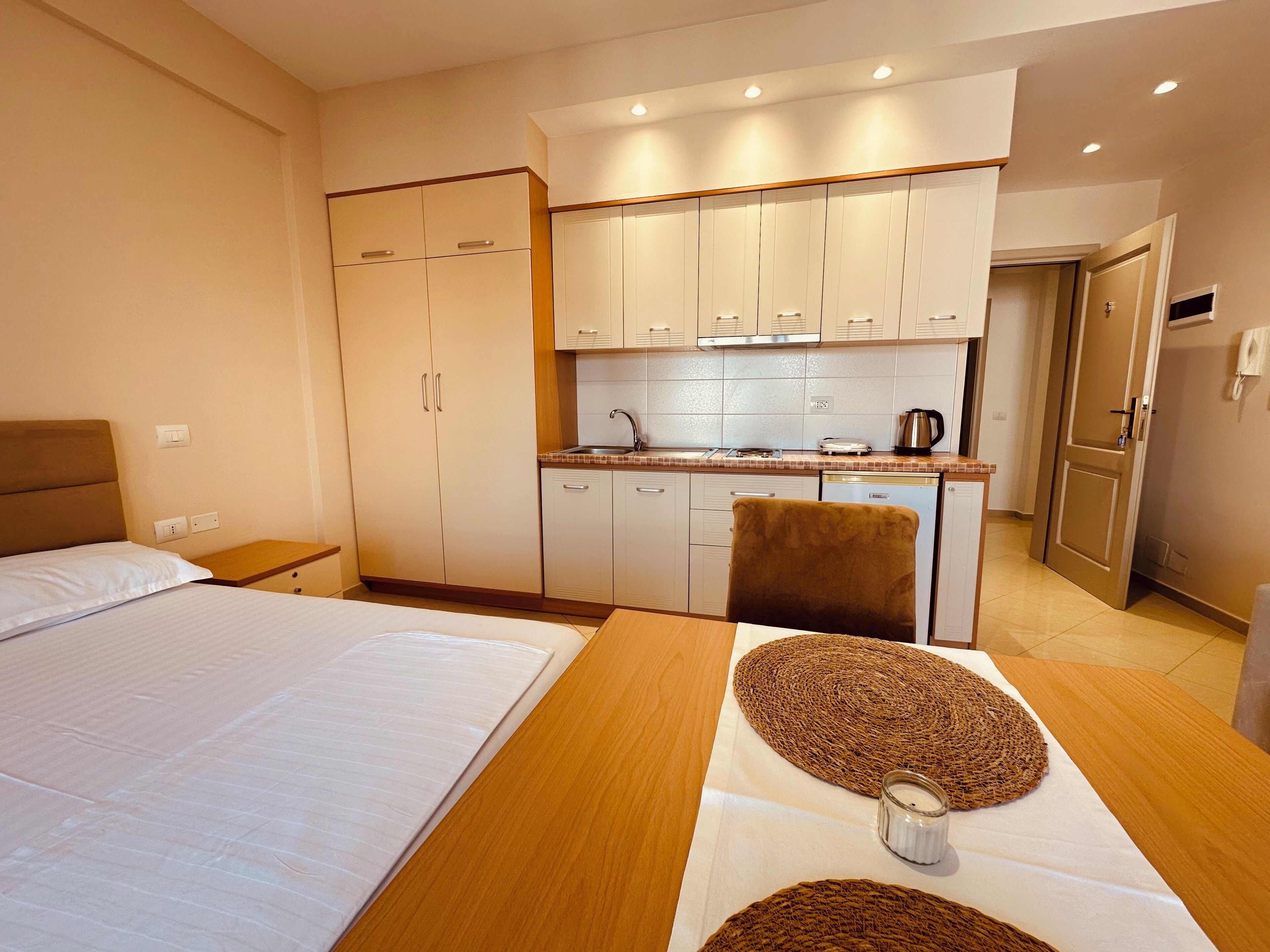 Quarto de hotel pequeno com cama, mesa de jantar com jogos americanos, área de cozinha com armários e porta de banheiro.