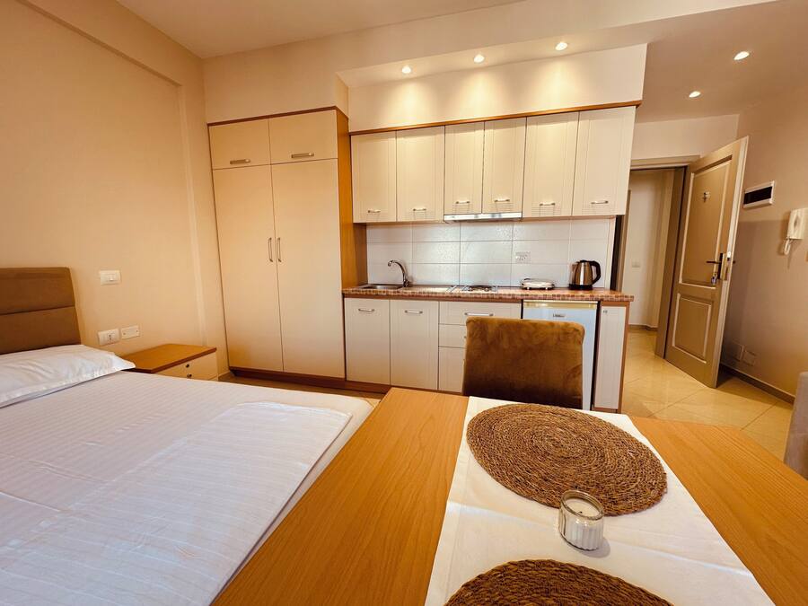 Quarto de hotel pequeno com cama, mesa de jantar com jogos americanos, área de cozinha com armários e porta de banheiro.