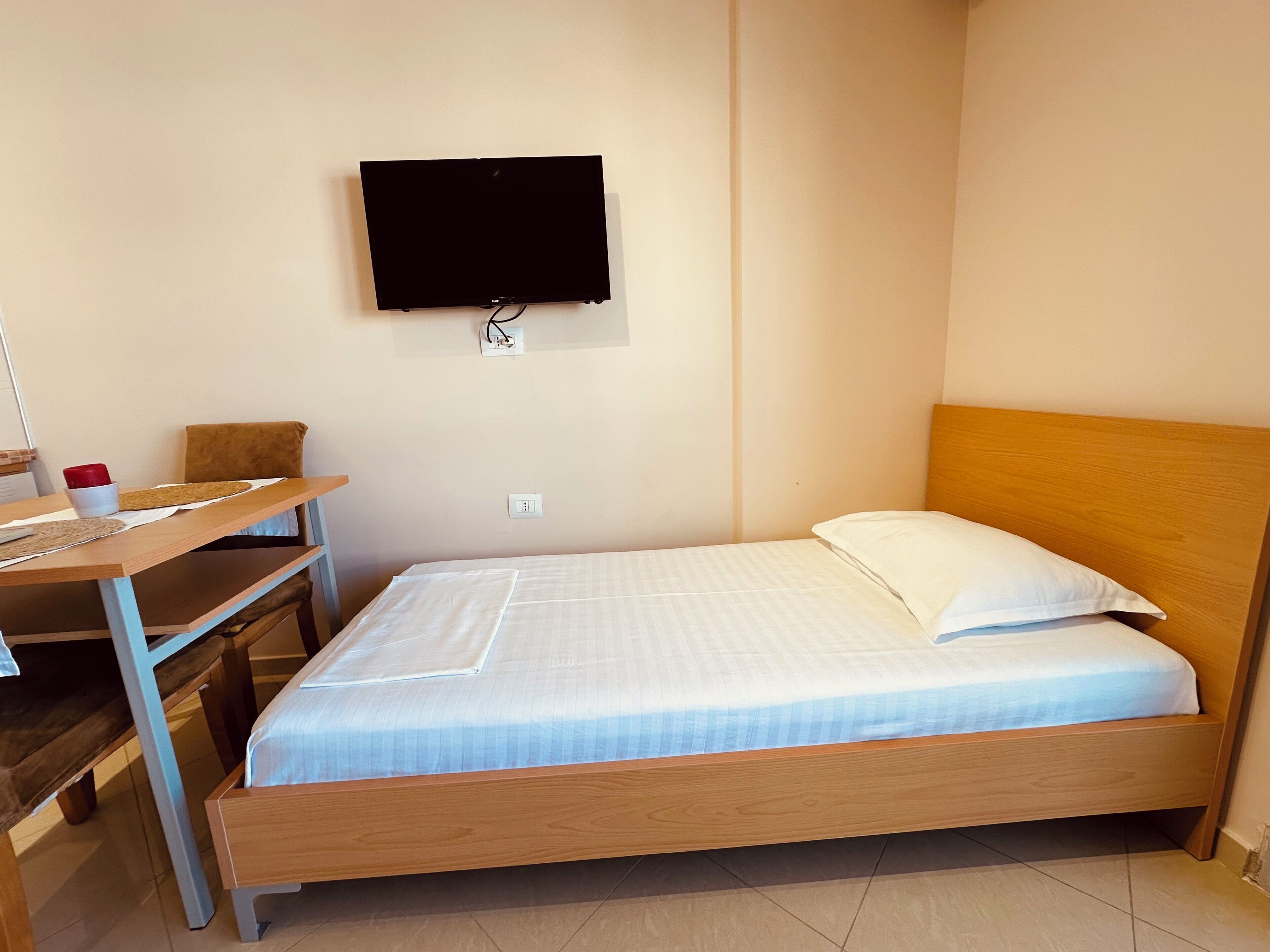 Quarto com cama de madeira, televisão de tela plana na parede, escrivaninha com cadeira e uma mesinha com uma xícara vermelha.