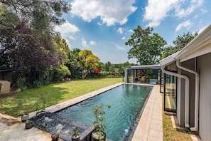 Uma piscina moderna ao ar livre com área de estar coberta por um terraço envidraçado, cercada por um gramado bem cuidado e diversas árvores.