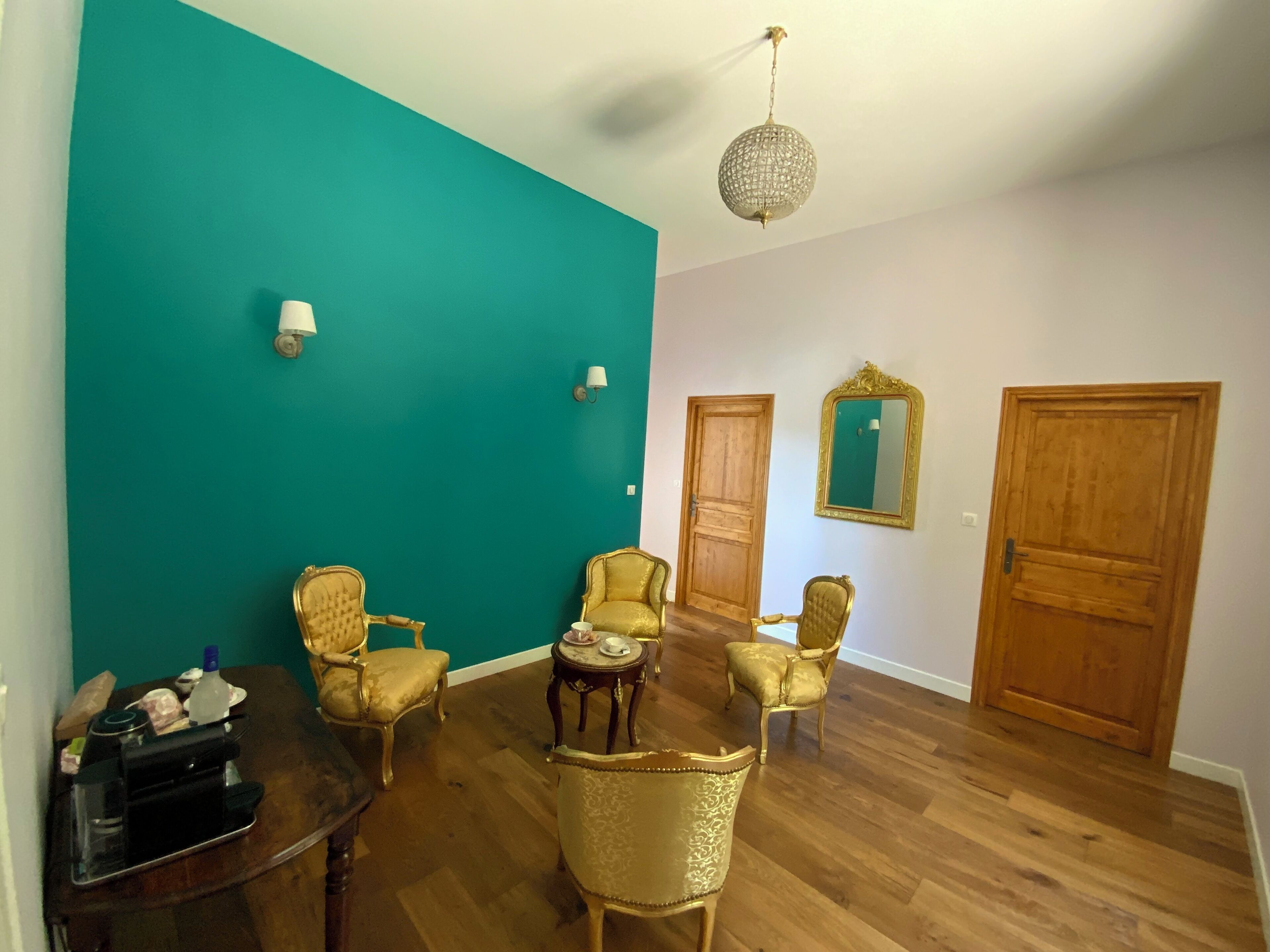 Quarto com paredes verde-água, piso de madeira e móveis com molduras douradas. Há duas portas, um espelho e um candelabro.