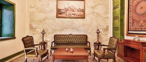 Um quarto com um sofá verde, duas poltronas, uma mesa de centro de madeira, um tapete estampado, um quadro na parede e uma porta verde.