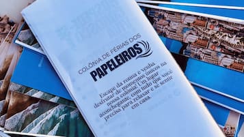 Um folheto intitulado 'Colônia de Férias dos Papelheiros' com uma ilustração de um horizonte urbano.