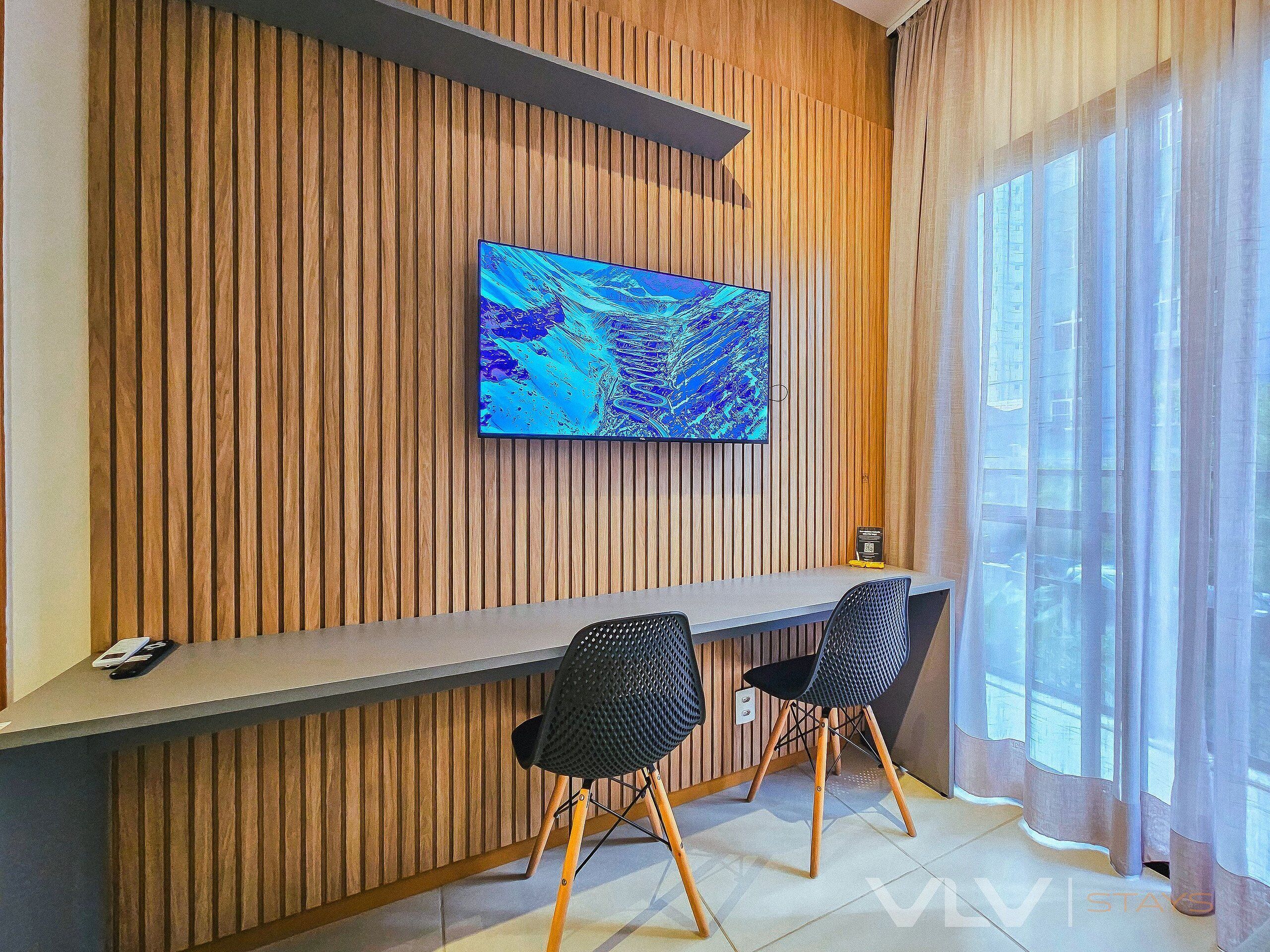 Quarto moderno com parede de painel de madeira, televisão de tela plana, escrivaninha, duas cadeiras e janela com cortinas.