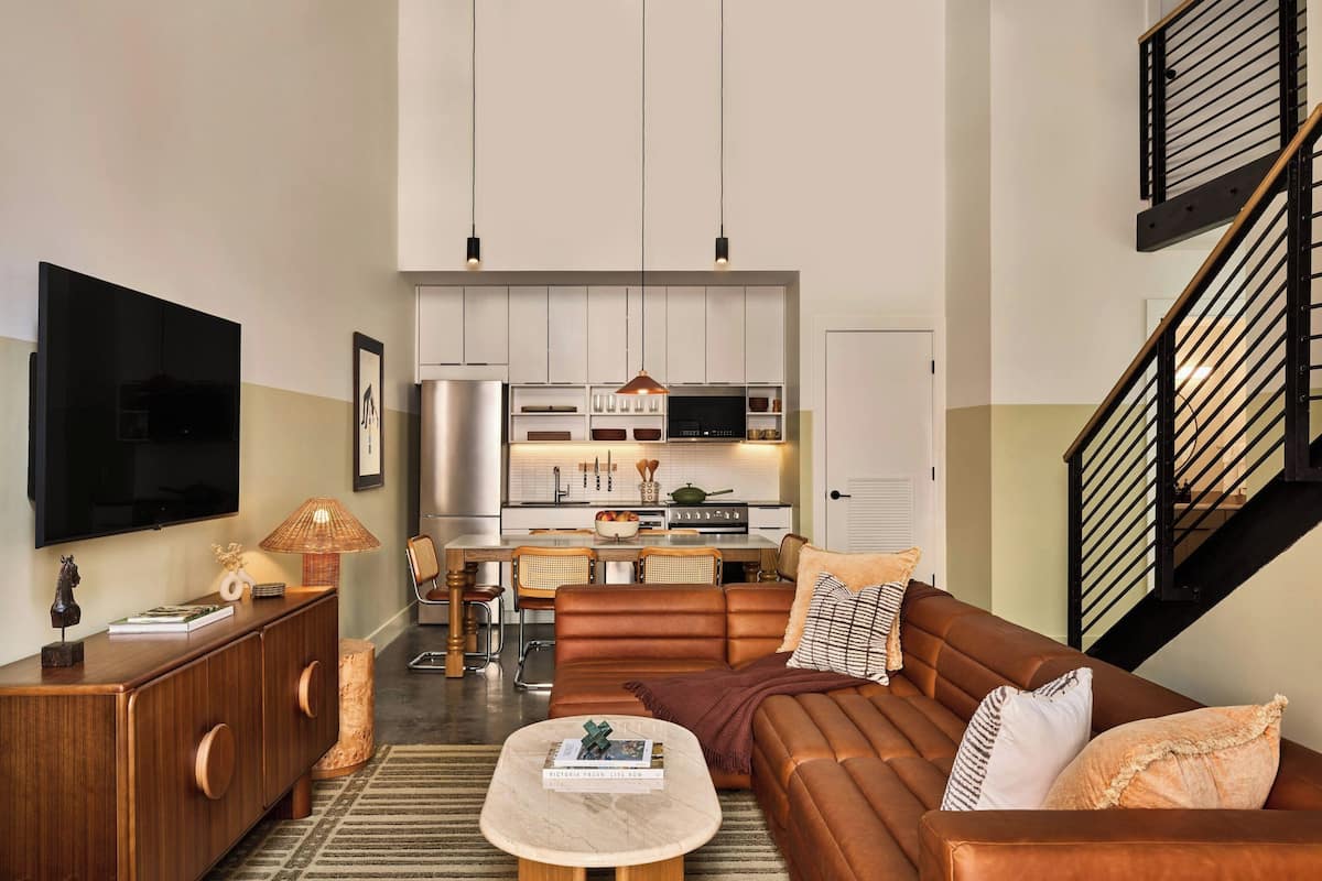 Uma sala de estar moderna com sofá, mesa de centro e televisão. Cozinha visível ao fundo, com armários e eletrodomésticos.