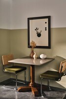 Uma sala de jantar com uma mesa, duas cadeiras, um vaso com flores secas e um quadro na parede.
