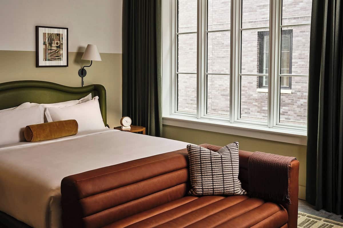 Quarto de hotel com cama, mesa de cabeceira, luminária, janela com cortinas e vista para um edifício de tijolos.