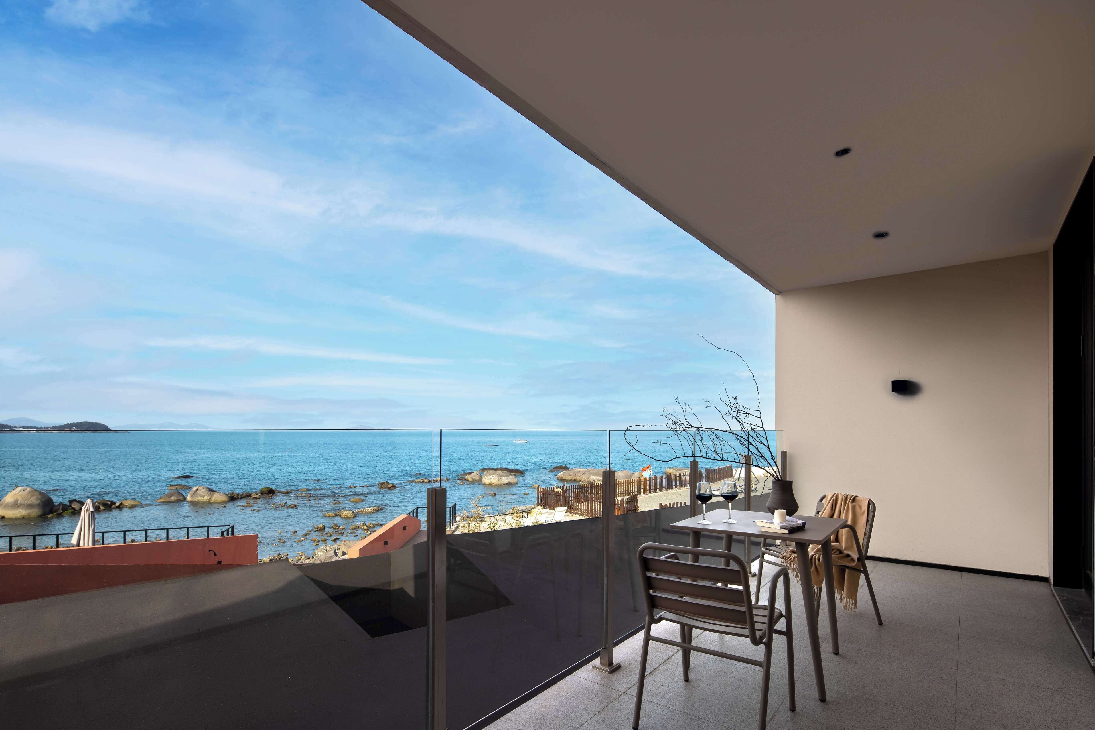 Uma varanda com vista para o mar, uma mesa com cadeiras e um painel de vidro.