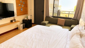 Quarto de hotel com cama, televisão, pequeno espaço de estar e janela com cortinas.