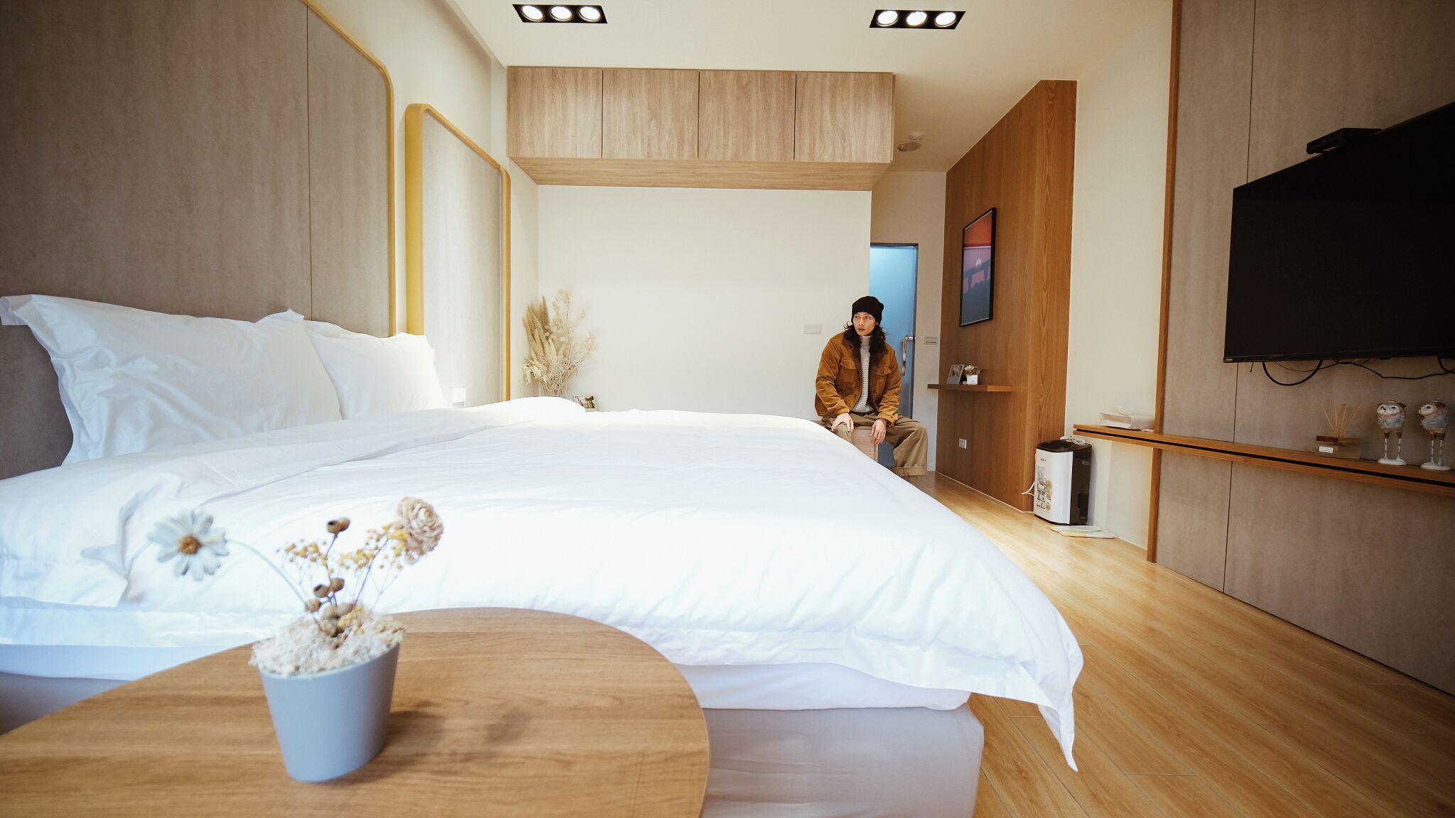 Quarto moderno com cama grande, mesa de cabeceira de madeira, TV e prateleira na parede.