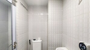 Casa de banho com paredes azulejadas em branco, sanita, lavatório e zona de duche.