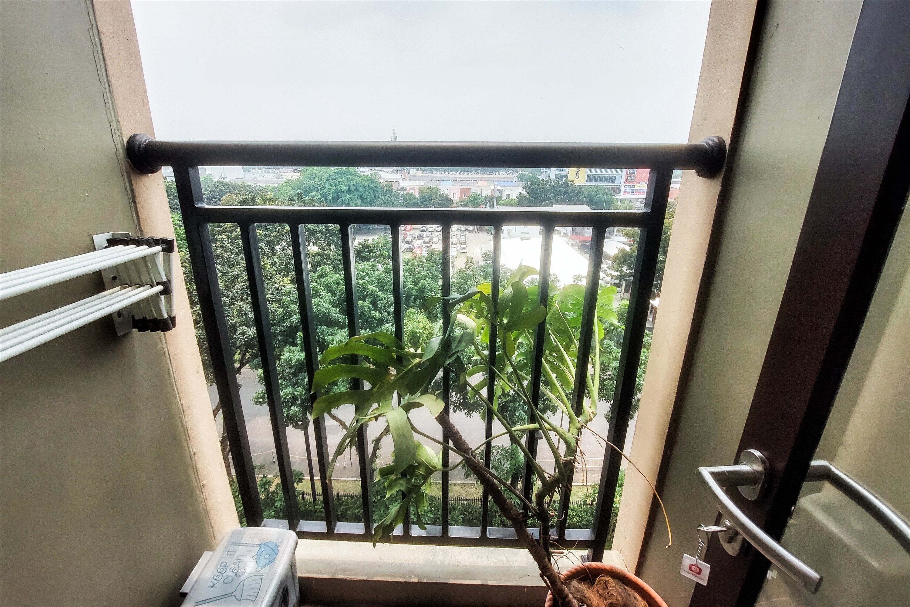 Varanda com grade de metal, um vaso de planta e vista para uma rua com edifícios e árvores.
