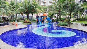 Piscina com design em mosaico azul, rodeada por palmeiras e um espaço de recreação infantil.