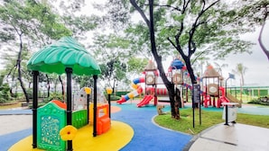 Um parquinho com estruturas coloridas para brincar, incluindo escorregadores e áreas de escalada, cercado por árvores e um caminho pavimentado.