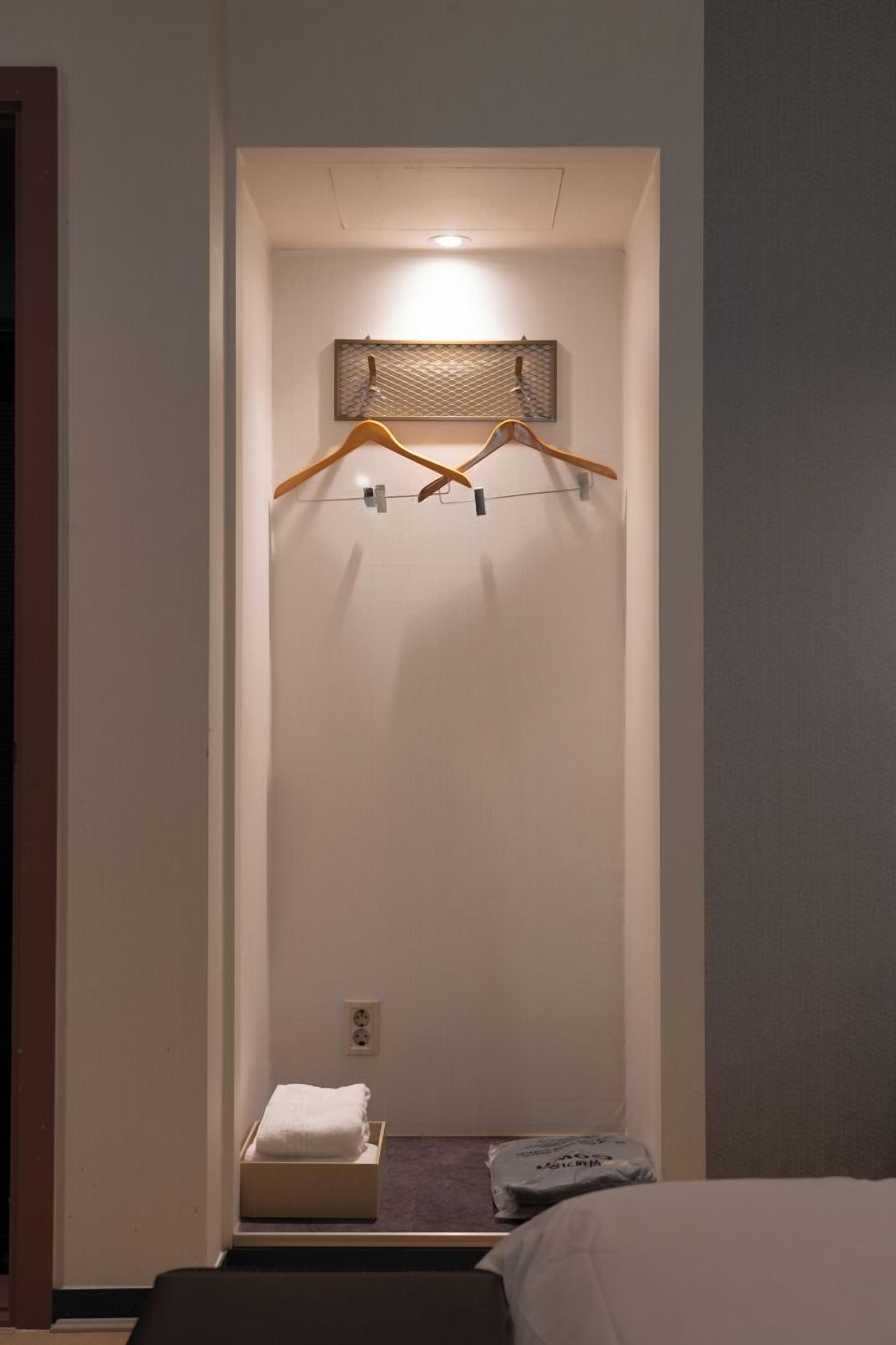 Quarto de hotel compacto com cama pequena, mesa de cabeceira, toalha e travesseiro na cama, um cabideiro com roupas e uma prateleira fixada na parede.