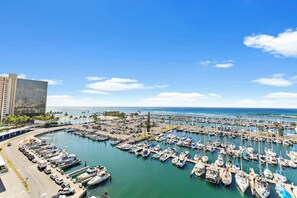 Uma marina com vários barcos atracados, céu azul e um edifício grande ao fundo.
