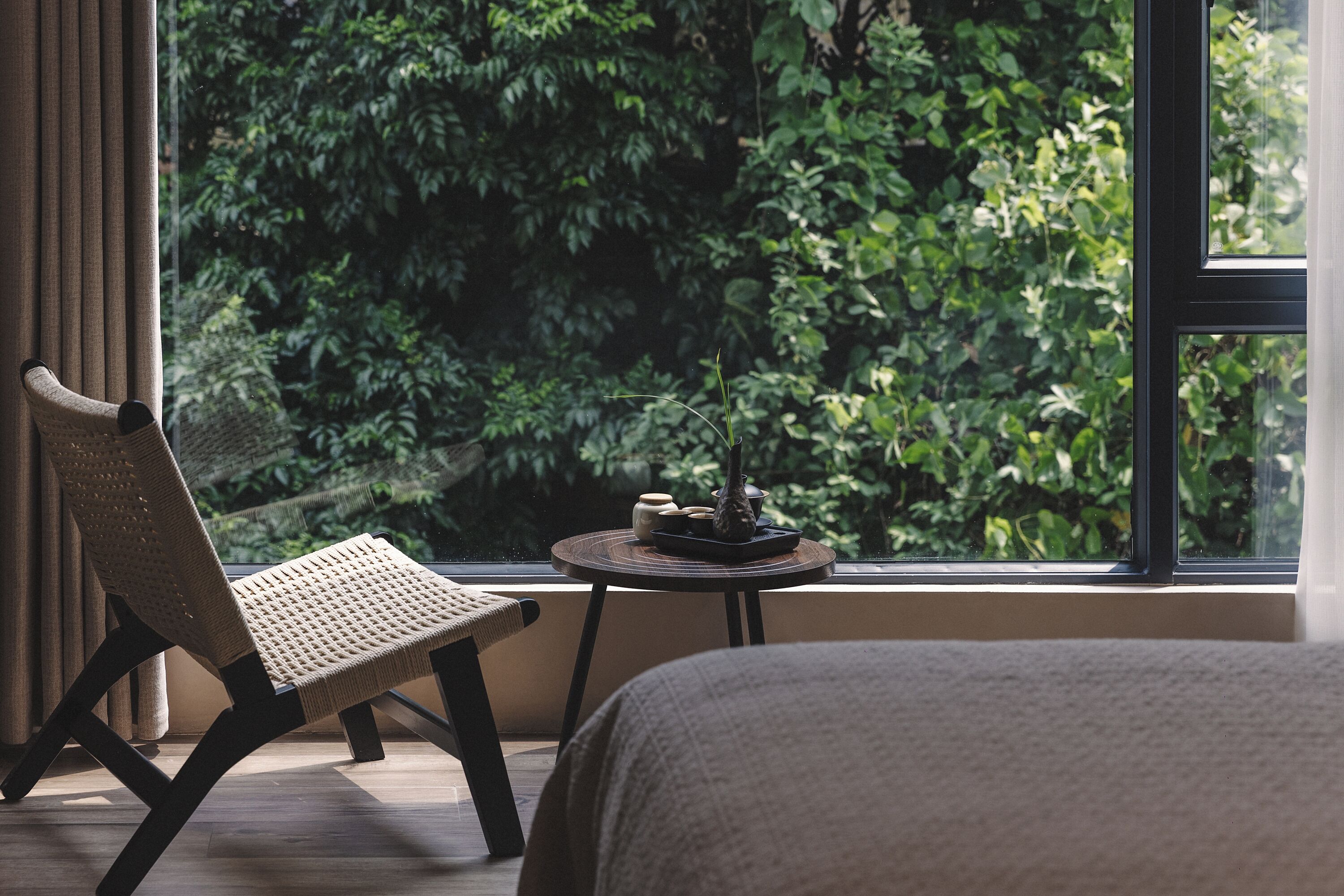 Quarto com uma cama, uma cadeira de vime, uma pequena mesa com uma planta e vista para um verde exuberante pela janela.